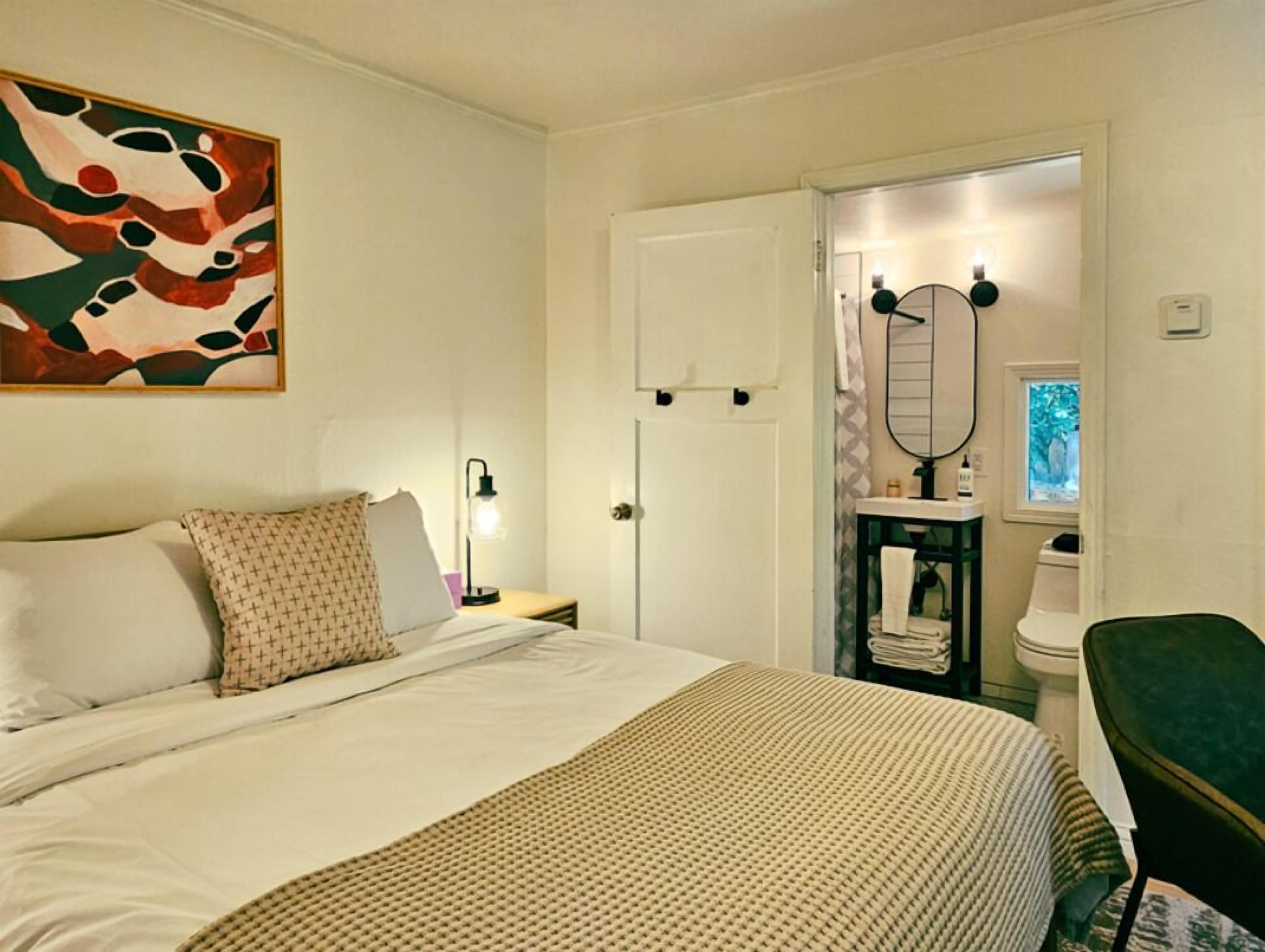 Quarto de hotel com cama, mesa de cabeceira, luminária, porta, espelho e uma cadeira.
