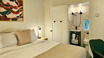 Quarto de hotel com cama, mesa de cabeceira, luminária, porta, espelho e uma cadeira.