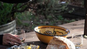 Uma pessoa servindo uma bebida em um copo, um prato com comida e uma tigela em uma mesa de madeira ao ar livre.