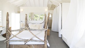 Uma cama de estilo rústico, com estrutura em madeira natural e roupa de cama branca, em um quarto claro e arejado, com teto alto e grandes janelas.