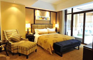 Quarto de hotel com uma cama grande, uma poltrona listrada, um banco, uma escrivaninha e uma janela ampla com vista.