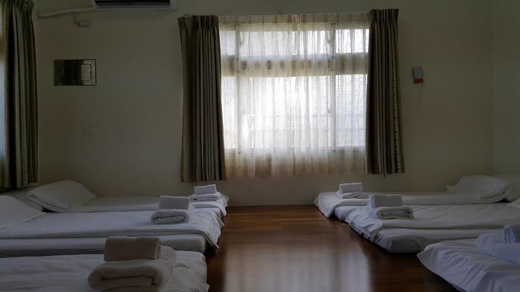 Quarto de hotel com quatro camas de solteiro dispostas em linha, piso de madeira e janela com cortinas leves.