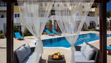Área ao lado da piscina com um sofá de vime, uma cesta de frutas e vista para a piscina e edifícios ao fundo.