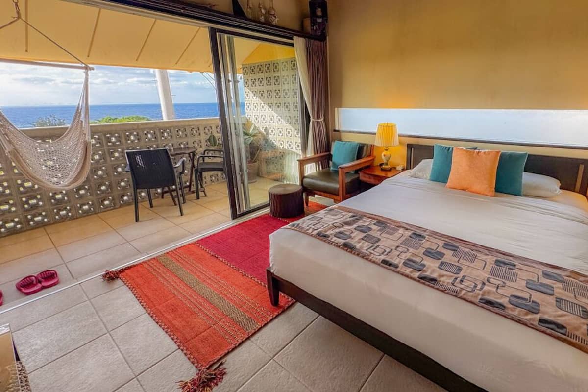 Quarto de hotel com cama, uma cadeira, mesinha, luminária e vista para o mar.