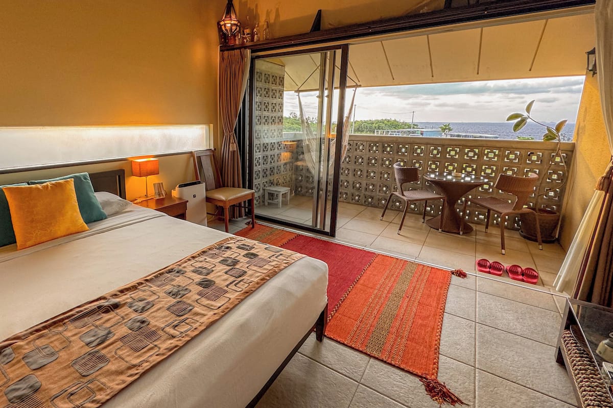 Quarto de hotel com uma cama grande, varanda com cadeiras e vista para o mar.