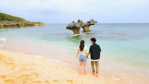 Um casal parado numa praia arenosa com água cristalina e uma formação rochosa ao longe.