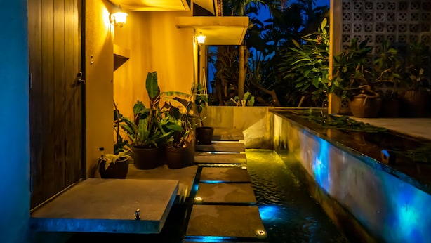 Vista noturna de um corredor de hotel com um elemento de água, plantas em vasos e iluminação quente.