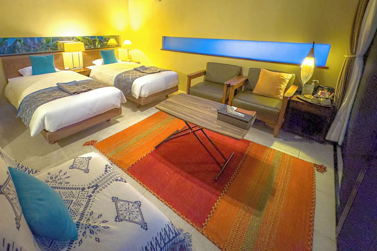 Quarto de hotel com duas camas, um sofá, uma mesa pequena e uma cadeira.