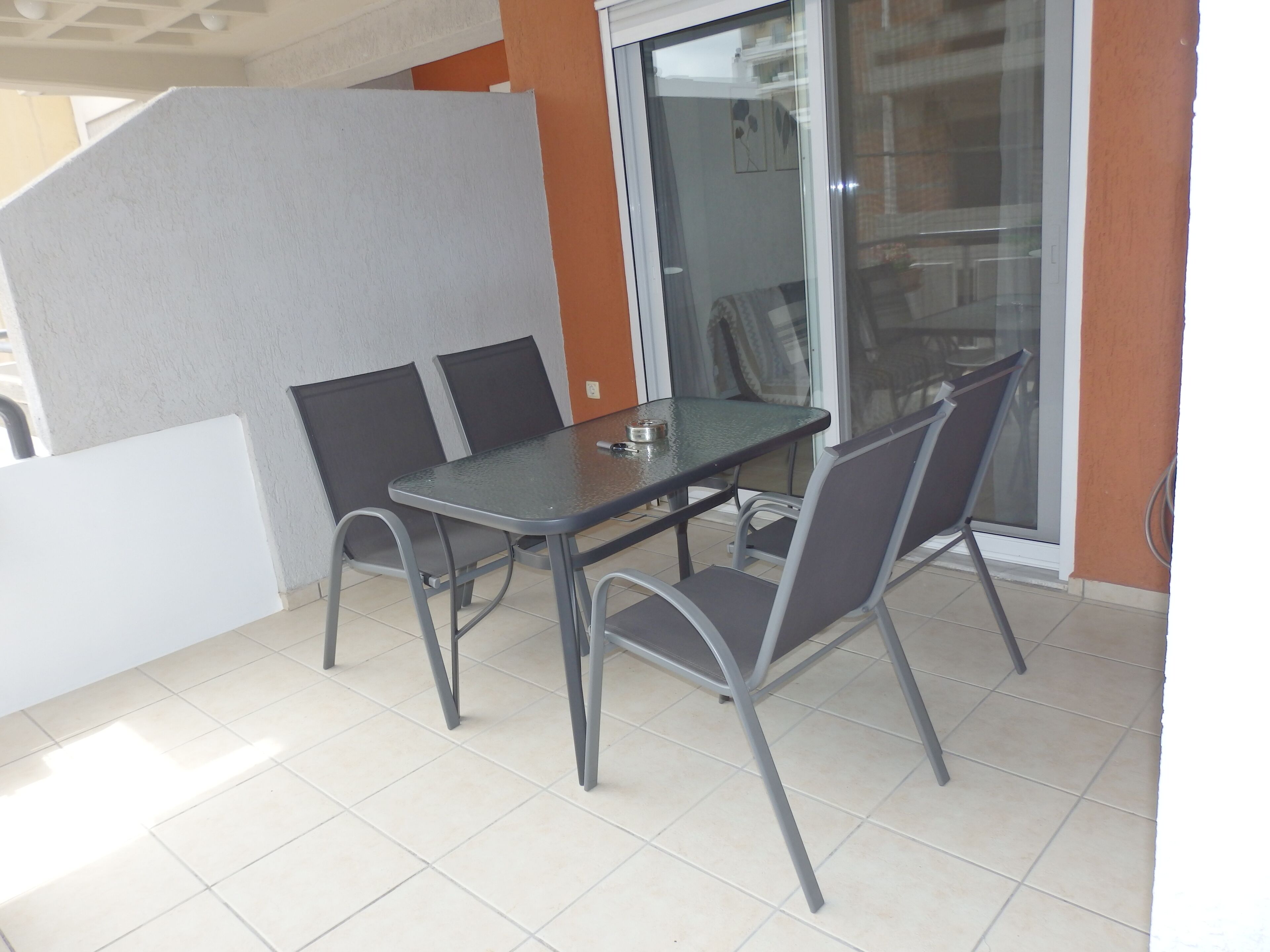 Uma mesa com tampo de vidro, quatro cadeiras, uma porta de correr em vidro e piso ladrilhado.