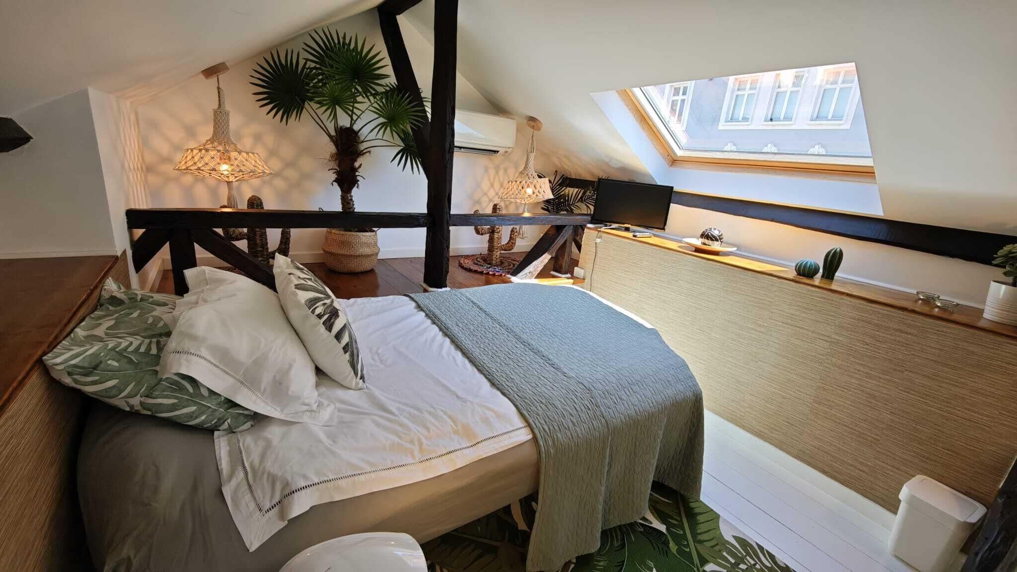 Quarto com cama, duas luminárias pendentes, uma mesinha e vista através de um claraboia.