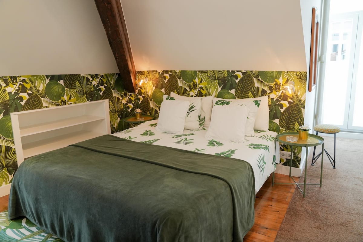 Quarto com cama, mesa de cabeceira, pequena mesa e uma cadeira. O quarto possui papel de parede com folhagem tropical e um vigamento de madeira no teto.