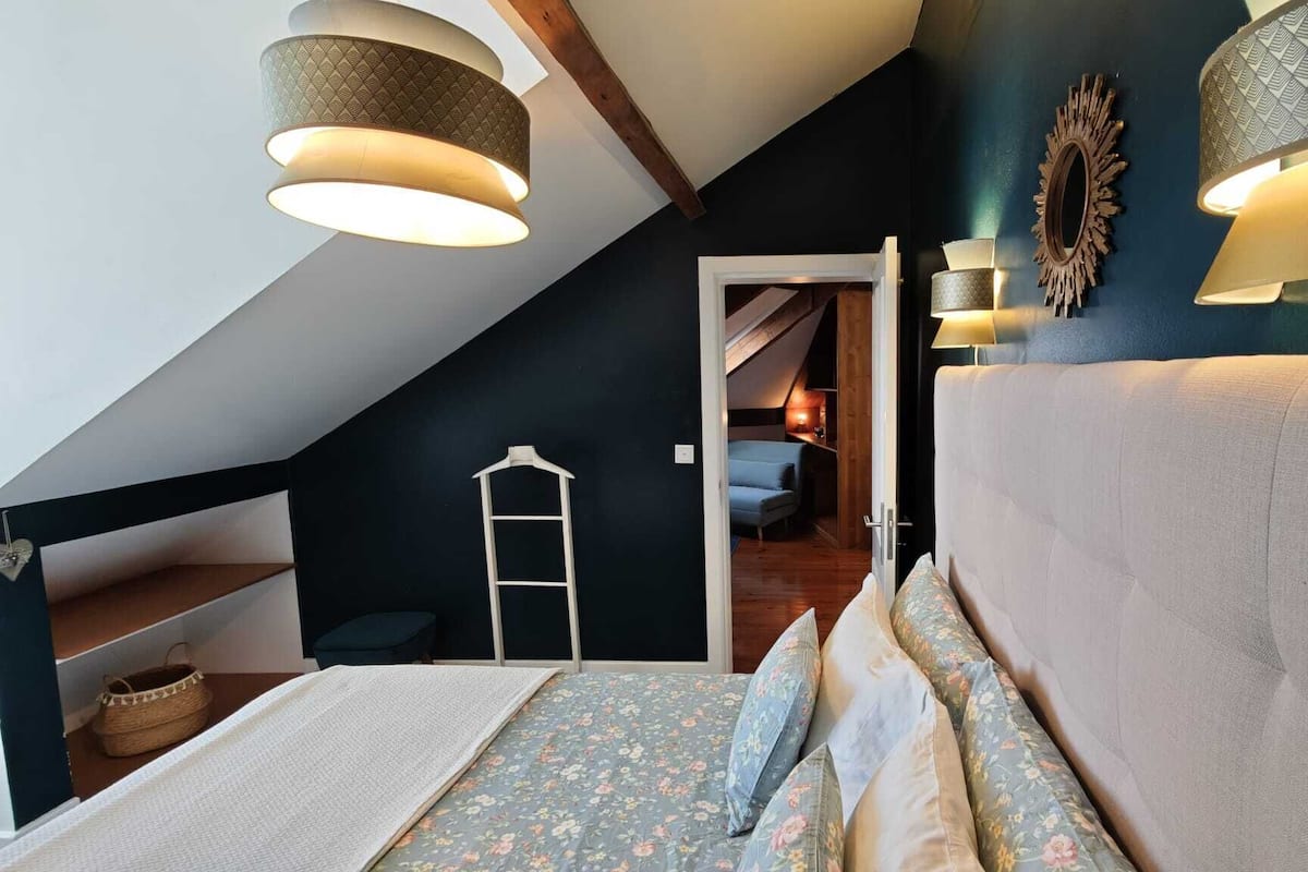 Um quarto de sótão aconchegante, com teto inclinado, uma cama com roupa de cama florida e duas luminárias fixadas na parede.