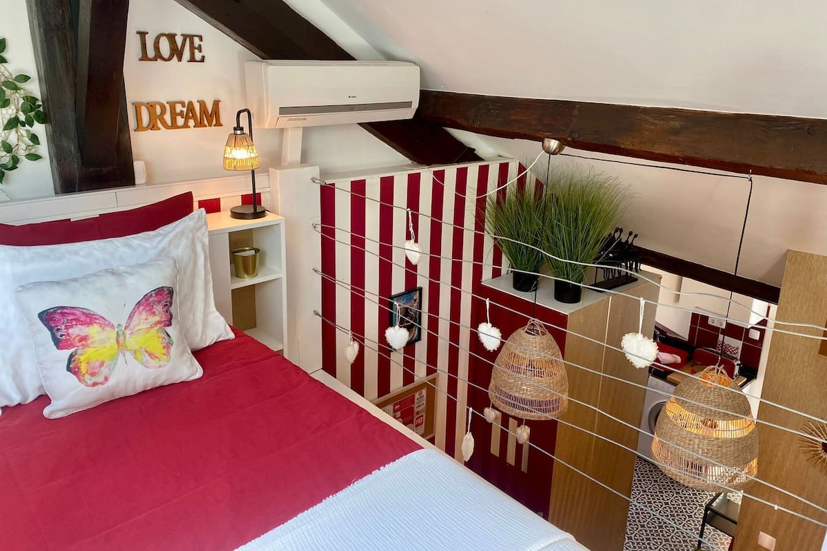 Quarto com cama, ar-condicionado fixado na parede, prateleira com objetos decorativos e um separador listrado em vermelho e branco.