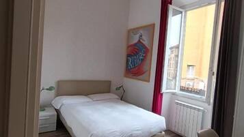 Quarto com cama, escrivaninha, cadeira, janela com cortinas e radiador.
