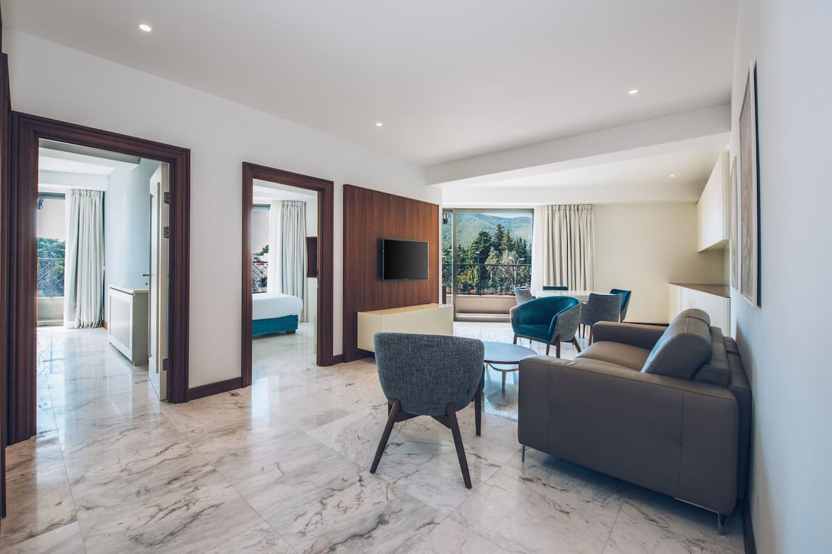 Uma sala de estar ampla com piso de mármore, um sofá, uma poltrona, uma TV fixada em um painel de madeira e uma varanda com vista para as montanhas.