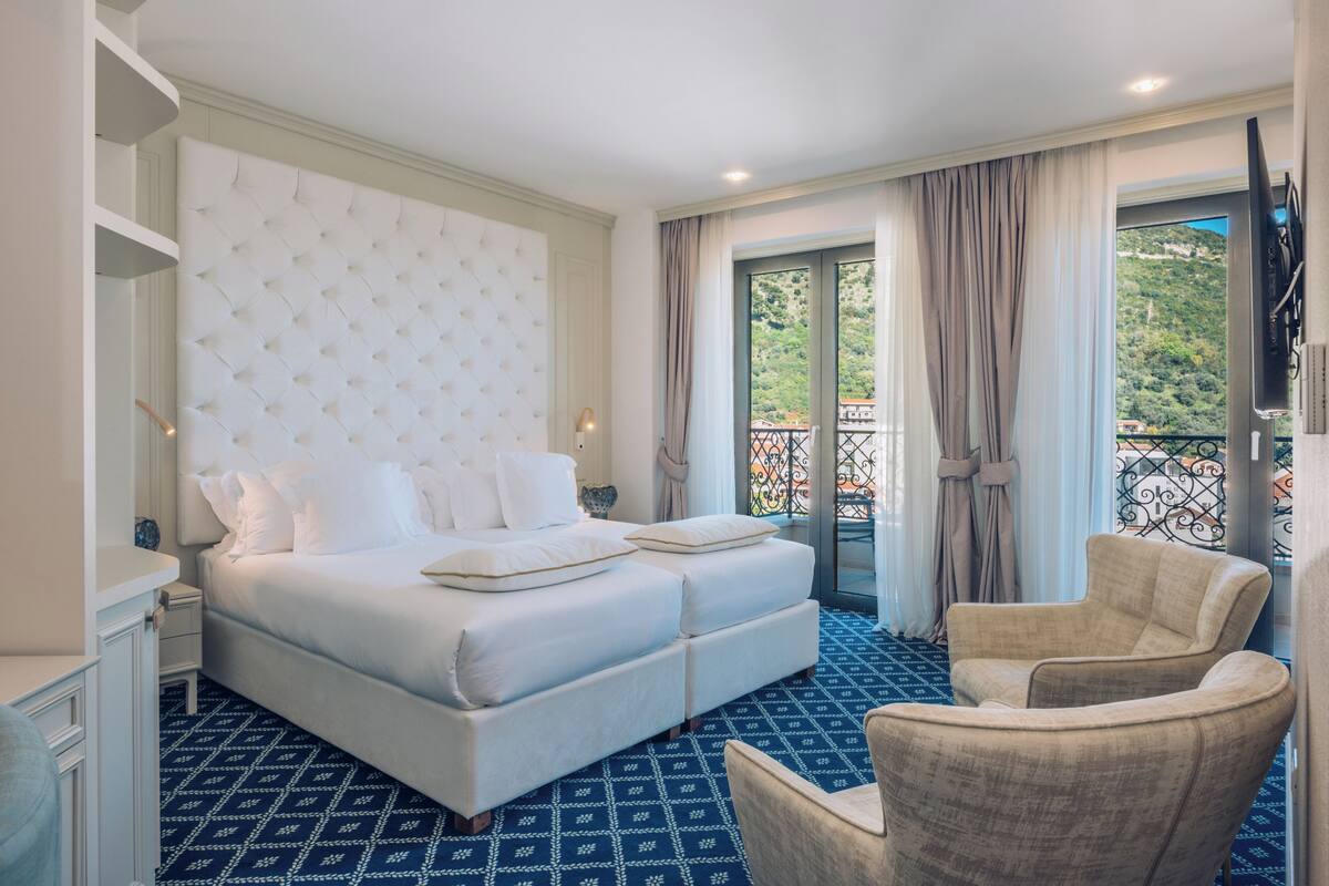 Quarto de hotel com uma cama grande, uma cadeira, uma mesinha de cabeceira e uma varanda com vista para o verde.
