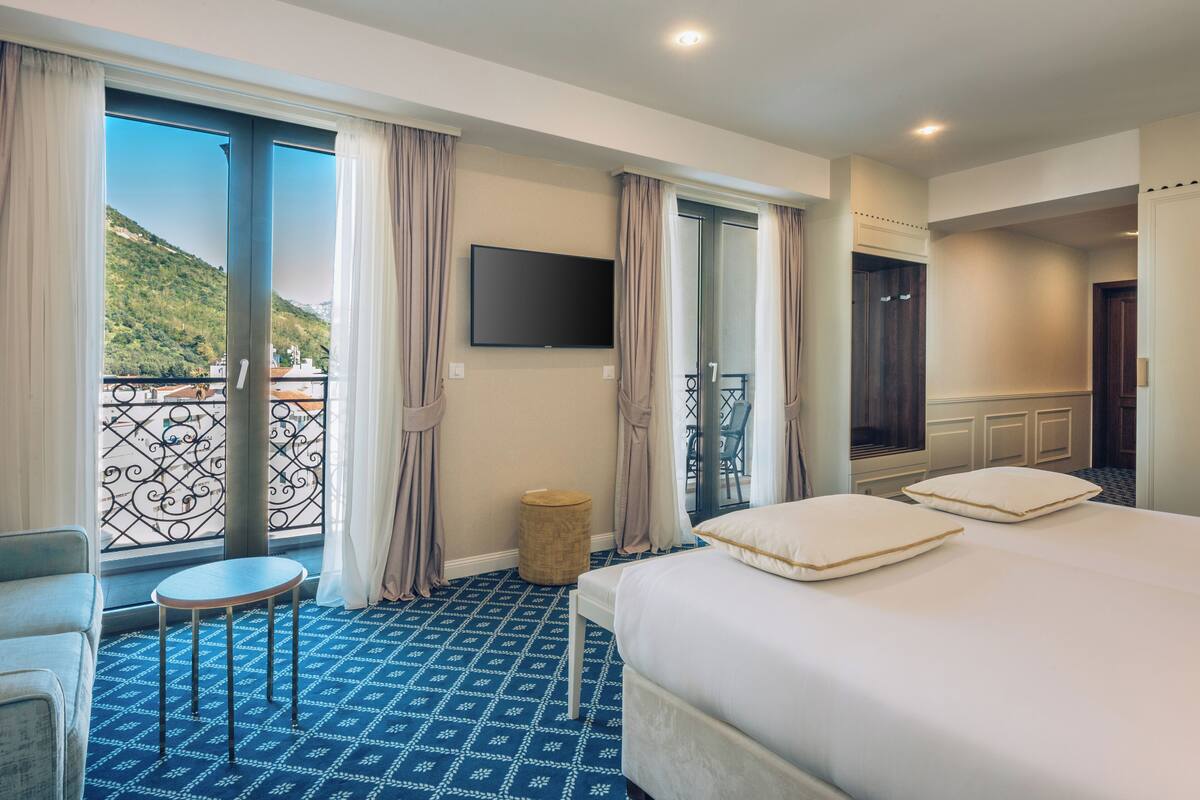 Quarto de hotel com uma cama grande, uma mesa pequena, televisão na parede e varanda com cortinas.