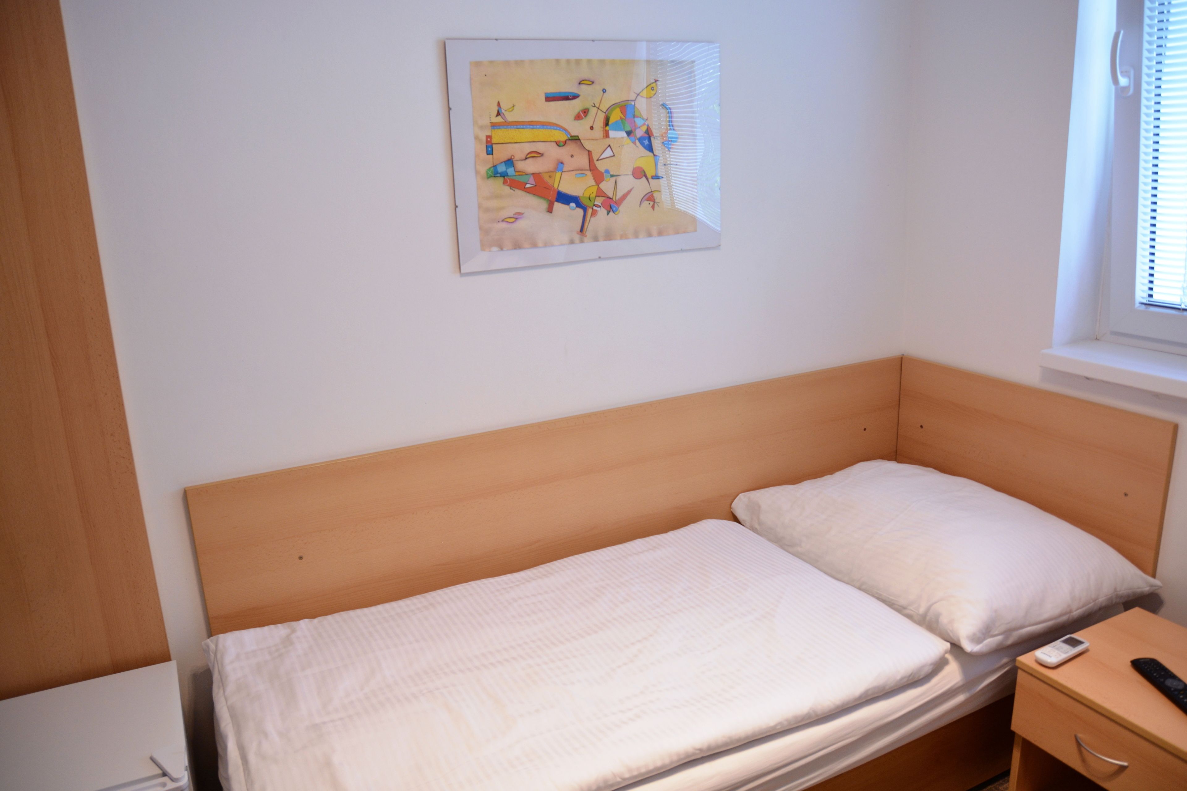 Uma cama de solteiro com cabeceira de madeira, uma mesa de cabeceira com controle remoto e uma janela com persianas.