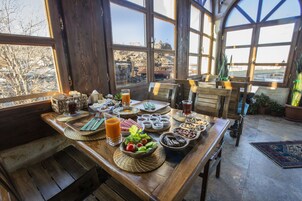 Mesa de jantar de madeira posta com diversos alimentos e bebidas, cercada por grandes janelas com vista para uma paisagem montanhosa.