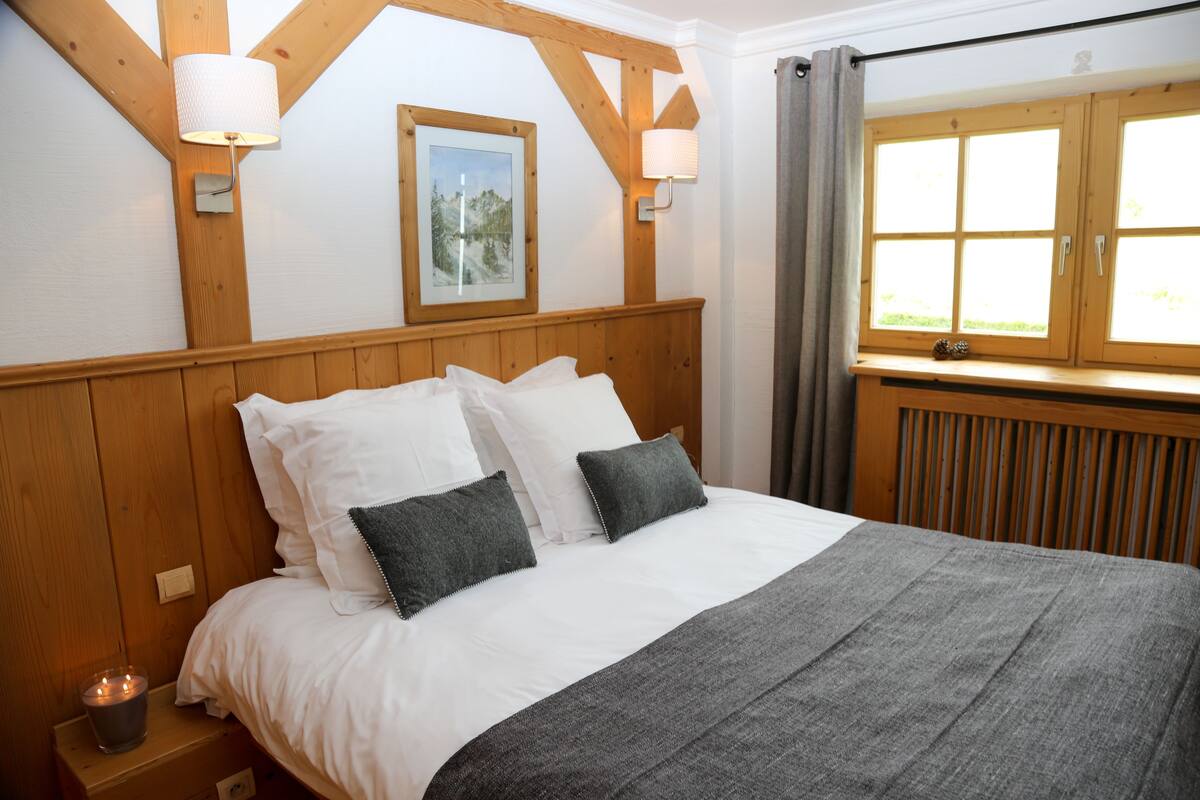 Cama bem arrumada com roupa de cama branca e cinza, cabeceira de madeira e um quadro na parede.