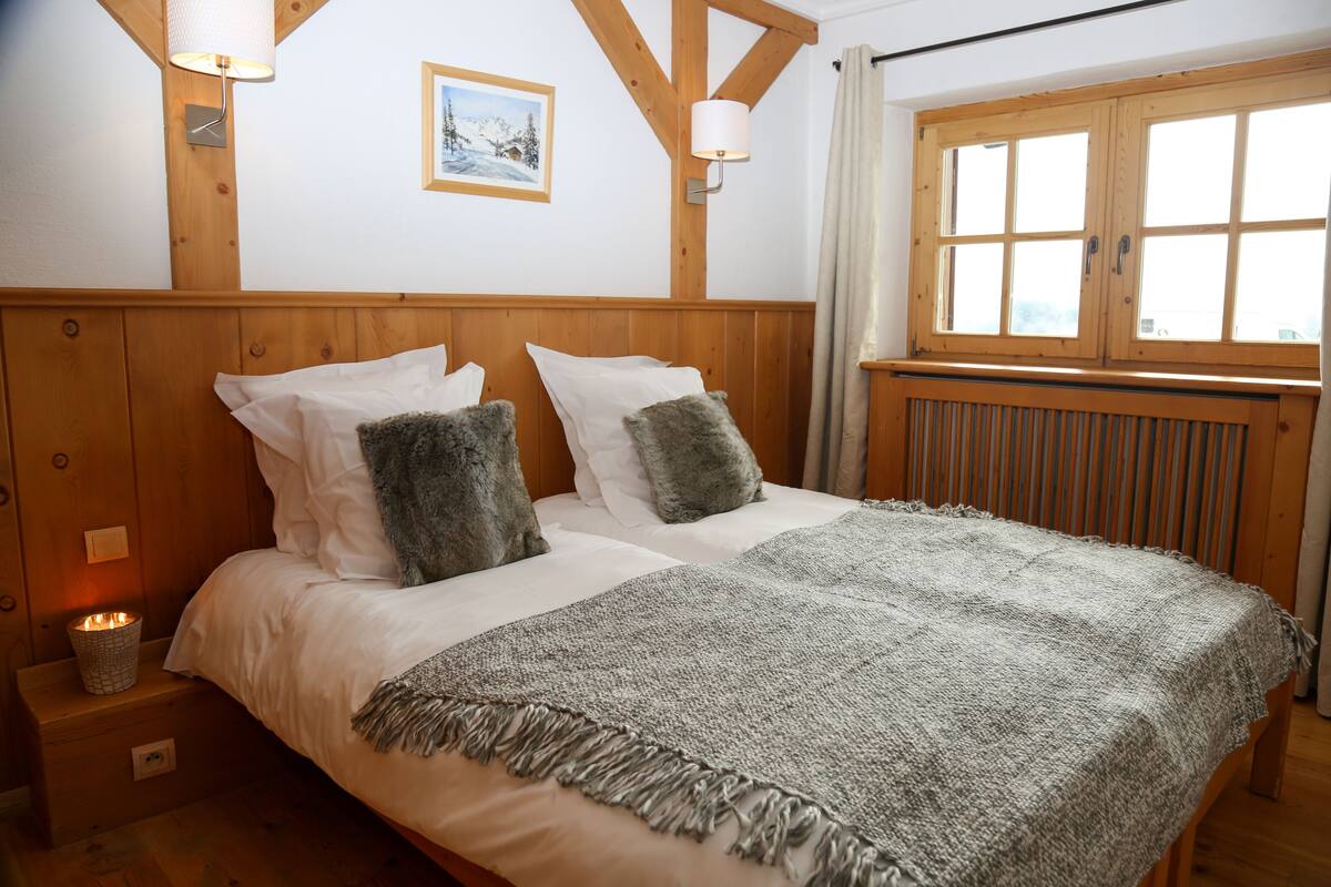 Quarto com cabeceira de madeira, cama grande com roupa de cama branca, duas almofadas de lançamento cinzas, manta cinza, abajur e janela com persianas de madeira.