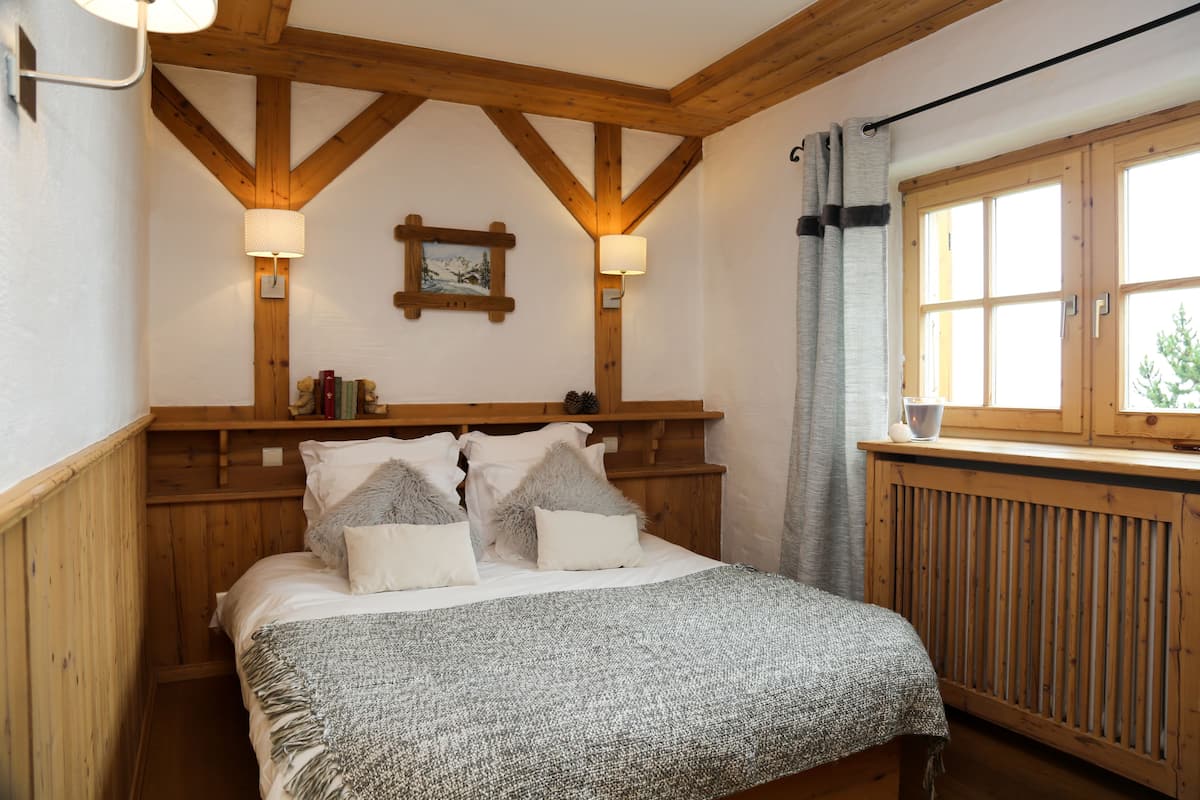 Quarto com cabeceira de madeira, duas luminárias laterais, um quadro emoldurado e janela com cortinas.