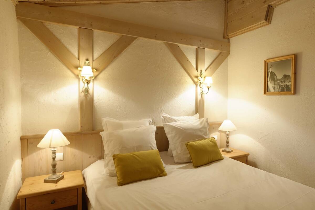 Quarto com teto de madeira, duas mesas de cabeceira com luminárias, cama com roupa de cama branca e dois travesseiros verdes, e um quadro na parede.