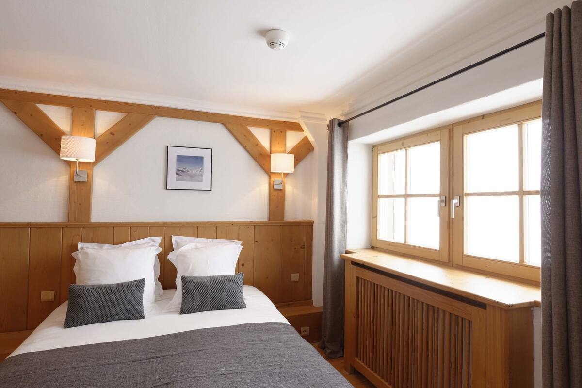 Quarto com cabeceira de madeira, uma janela grande com cortinas e um radiador.
