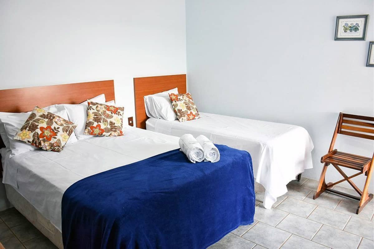 Quarto de hotel com duas camas de solteiro, cada uma com cabeceira de madeira, roupa de cama branca e almofadas com padrão floral. Um cobertor azul está dobrado sobre a cama à esquerda, e dois toalhas enroladas sobre a cama à direita. Uma cadeira de madeira está posicionada à direita das camas.