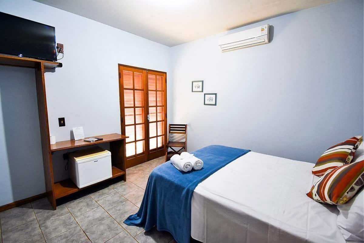 Quarto de hotel compacto com uma cama, uma mesa com computador, uma cadeira e um pequeno frigorífico.
