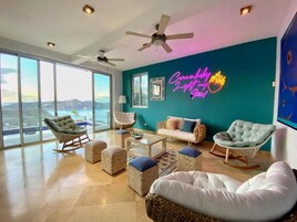 Uma sala de estar espaçosa com uma grande janela, móveis de vime, um letreiro de neon e ventiladores de teto.