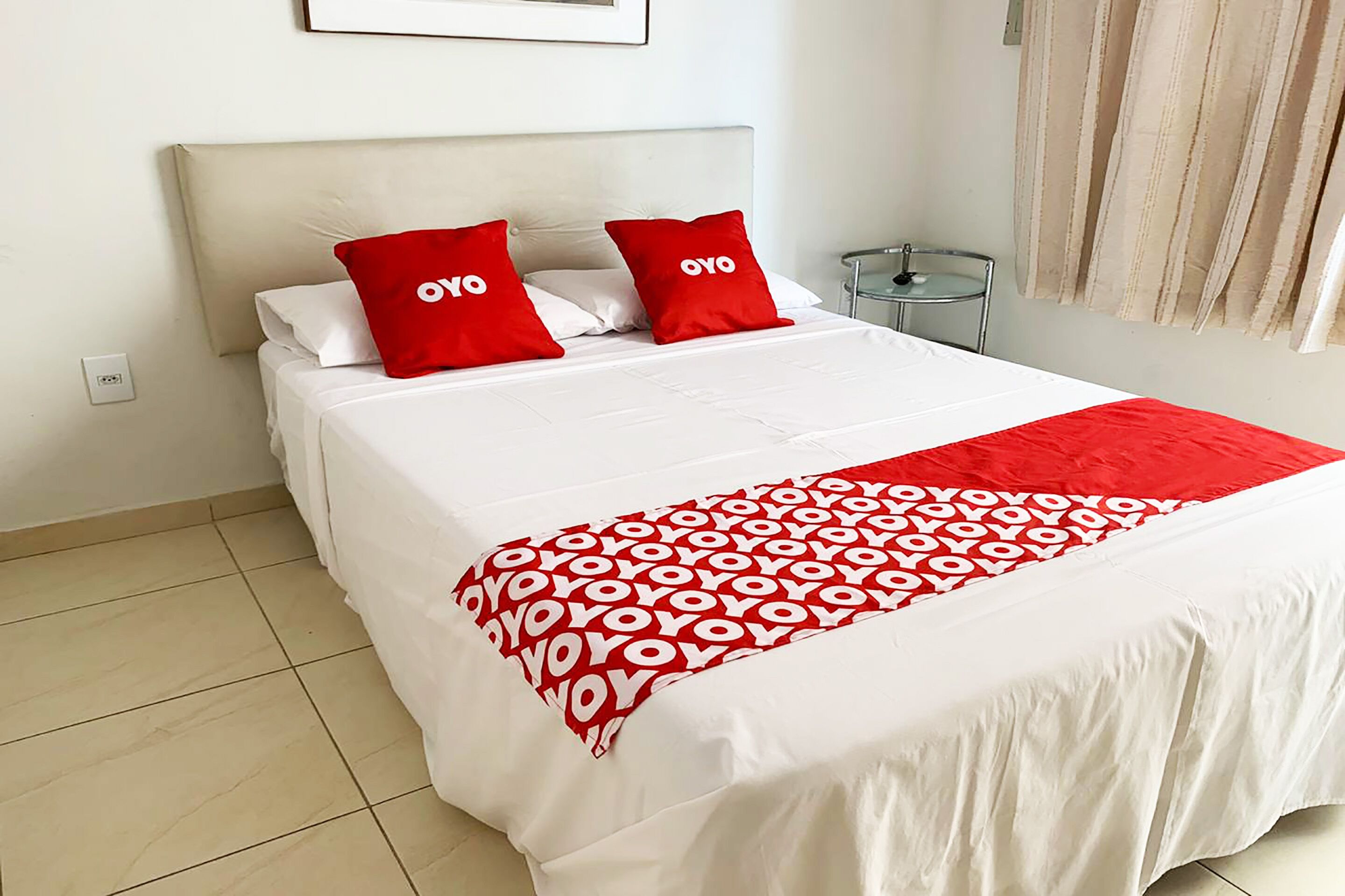 Quarto de hotel com cama bem arrumada, duas almofadas vermelhas com a marca 'OYO', colcha branca e manta estampada em vermelho e branco.