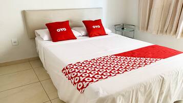 Quarto de hotel com cama bem arrumada, duas almofadas vermelhas com a marca 'OYO', colcha branca e manta estampada em vermelho e branco.