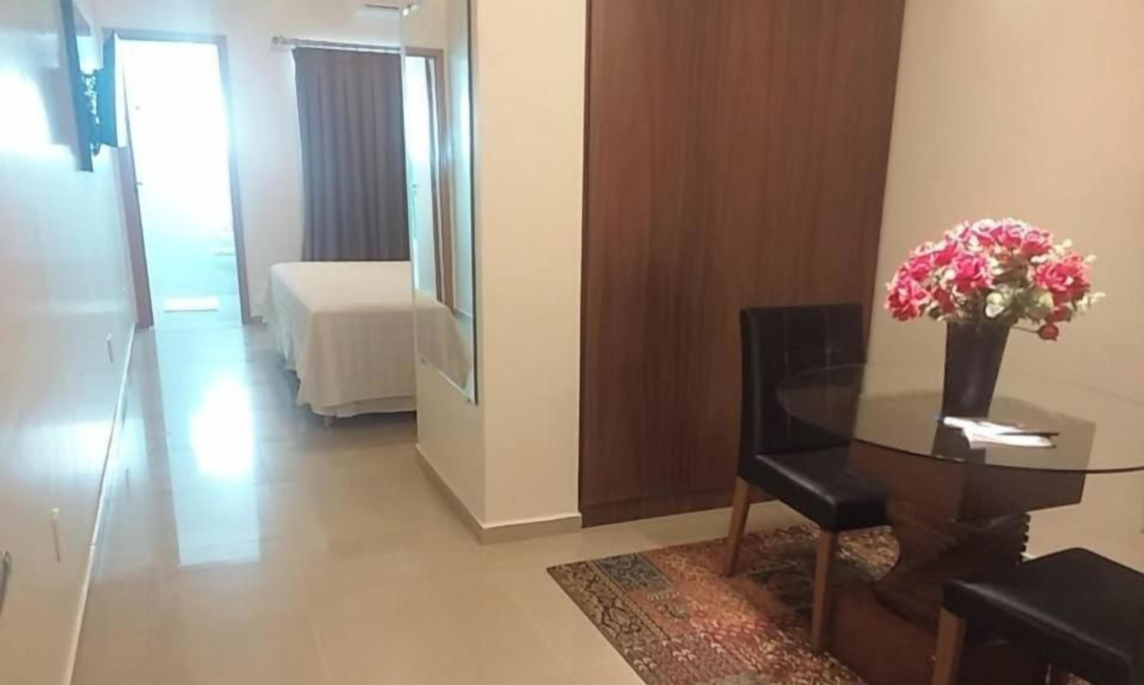 Quarto de hotel com cama, uma pequena mesa com cadeiras, um vaso de flores e vista para o corredor.