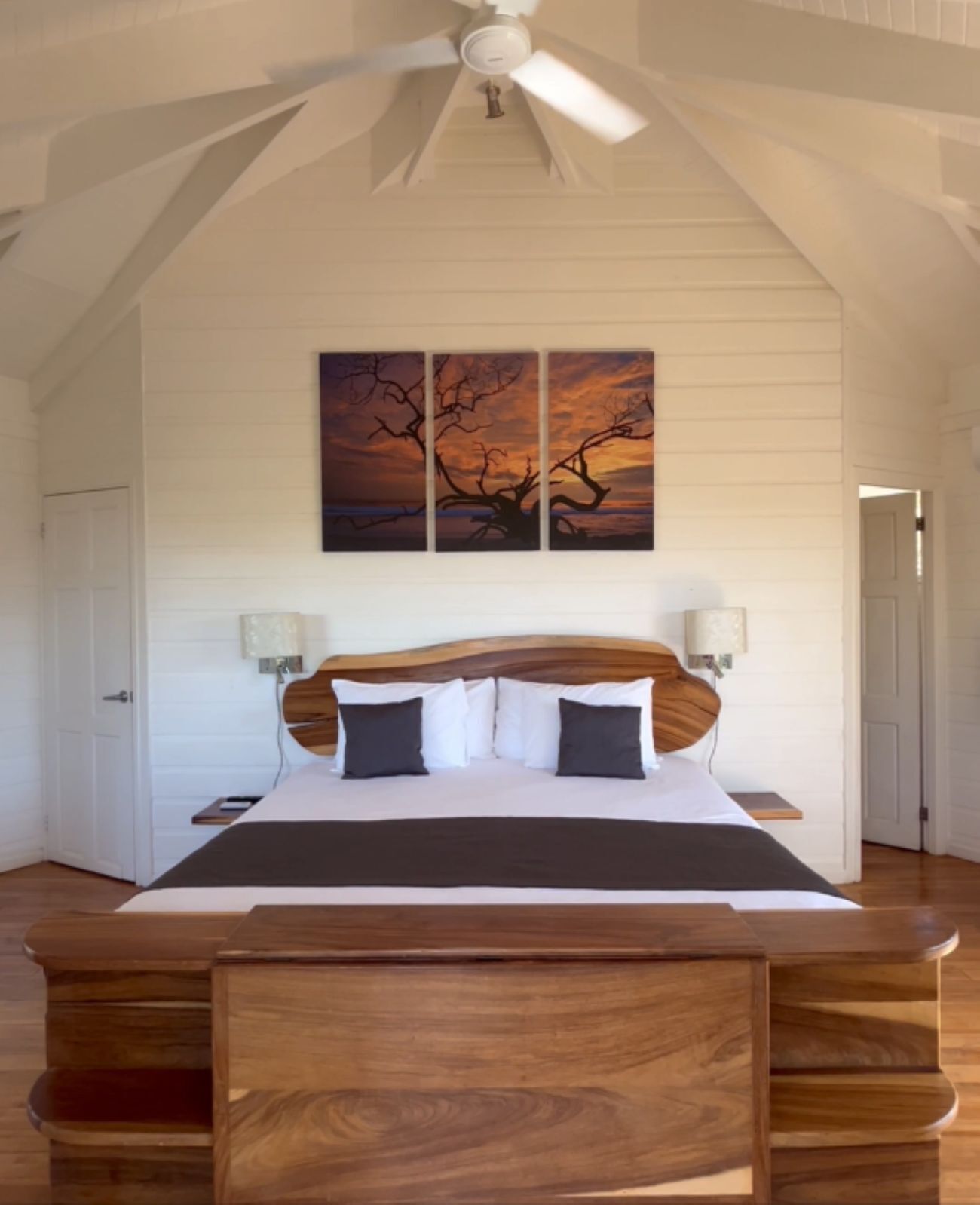Quarto espaçoso com uma cama grande, cabeceira de madeira e ventilador de teto. Há duas luminárias na parede e uma pintura com a silhueta de uma árvore sobre a cama.