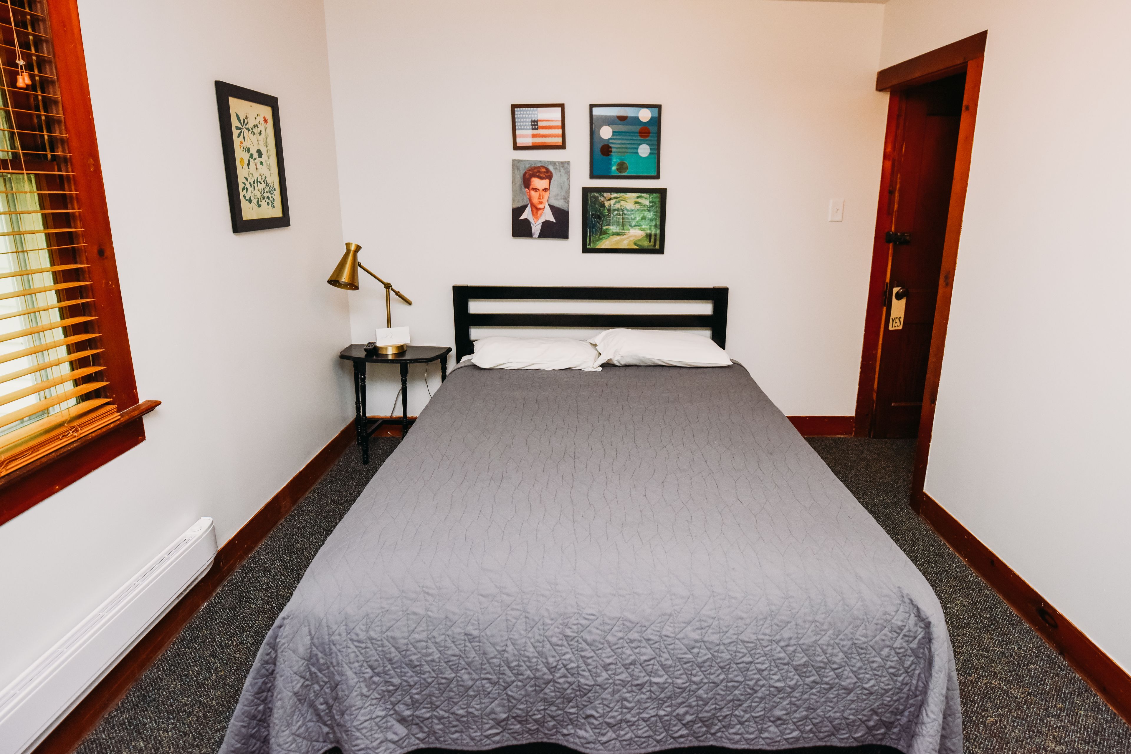 Cama bem arrumada num quarto de hotel, com cabeceira, mesa de cabeceira com abajur e quadros nas paredes.