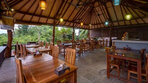 Um restaurante ao ar livre com mesas e cadeiras de madeira, telhado de palha e lanternas penduradas.