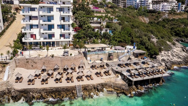 Vista aérea de um hotel à beira-mar, com vários andares, área de lazer na praia com espreguiçadeiras e guarda-sóis, e um litoral rochoso.