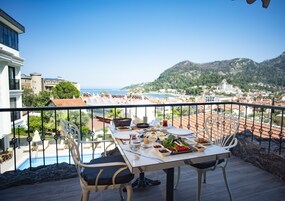 Terraço com mesa posta para refeição, com vista para uma cidade costeira e montanhas.