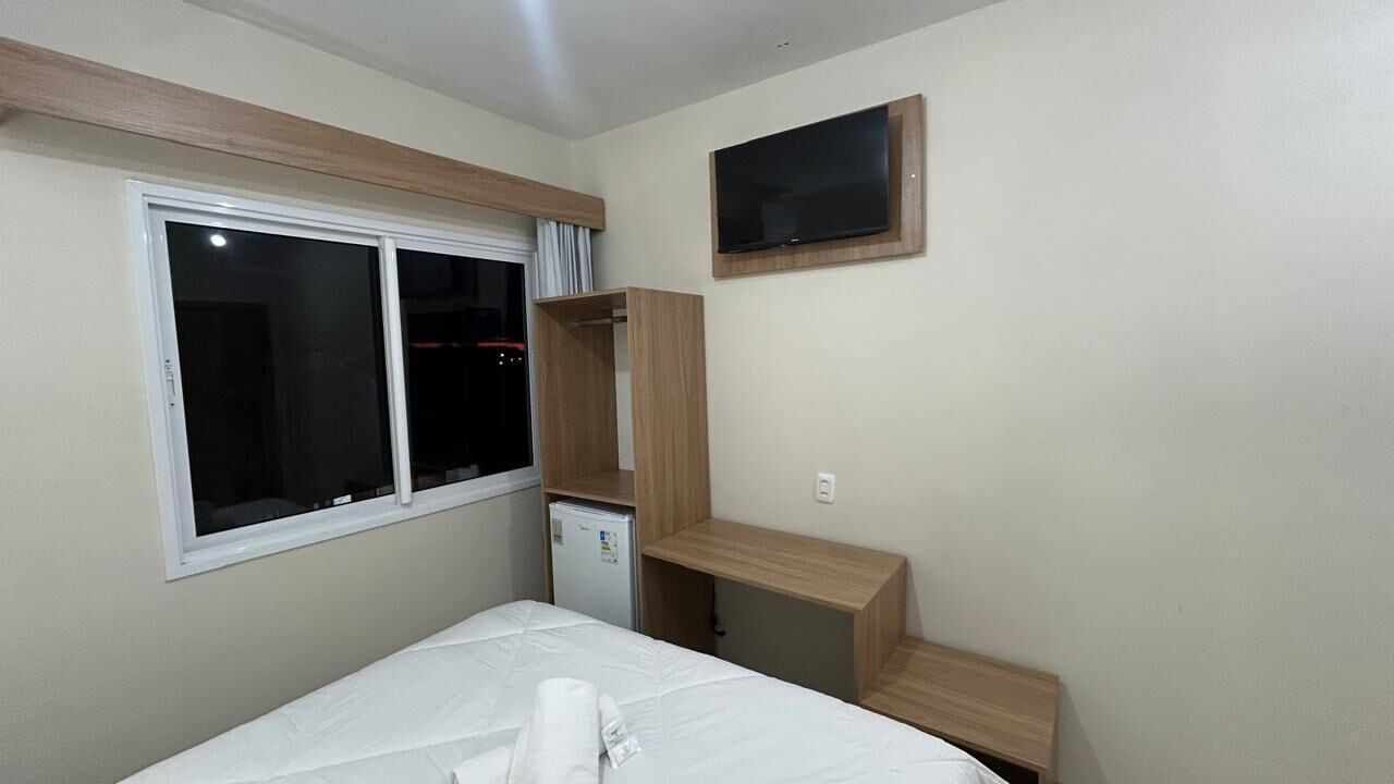 Quarto de hotel pequeno e funcional, com cama, escrivaninha, televisão presa à parede e janela com cortinas.