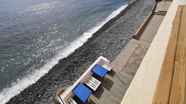 Uma praia com costa rochosa, um deck de madeira com assentos e vista para o oceano.
