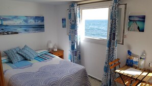 Um quarto com cama, mesa de cabeceira, uma mesa com cadeira, janela com cortinas e um quadro na parede.