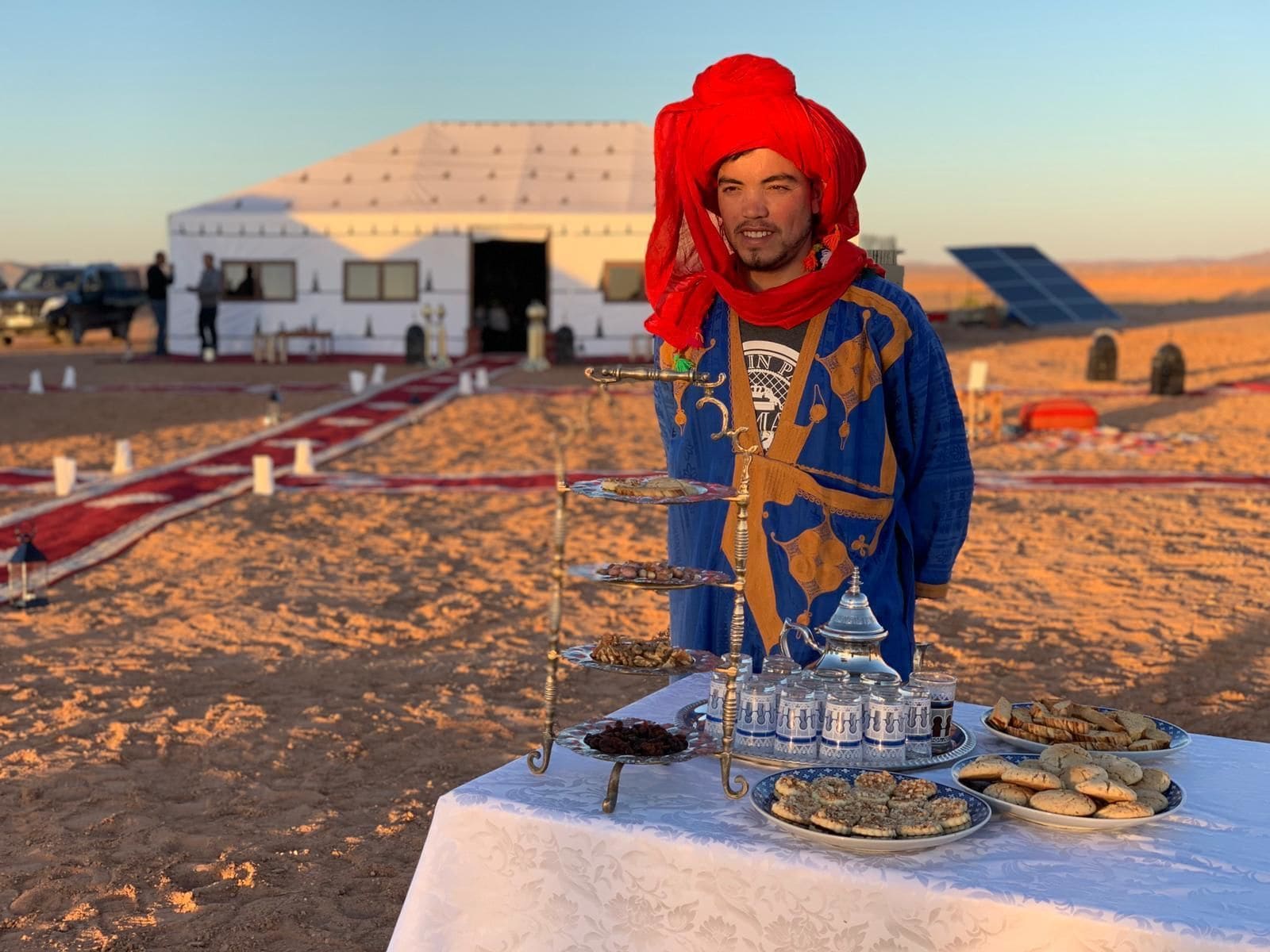 Uma pessoa vestida com traje tradicional em frente a uma mesa com comida, com uma tenda ao fundo.