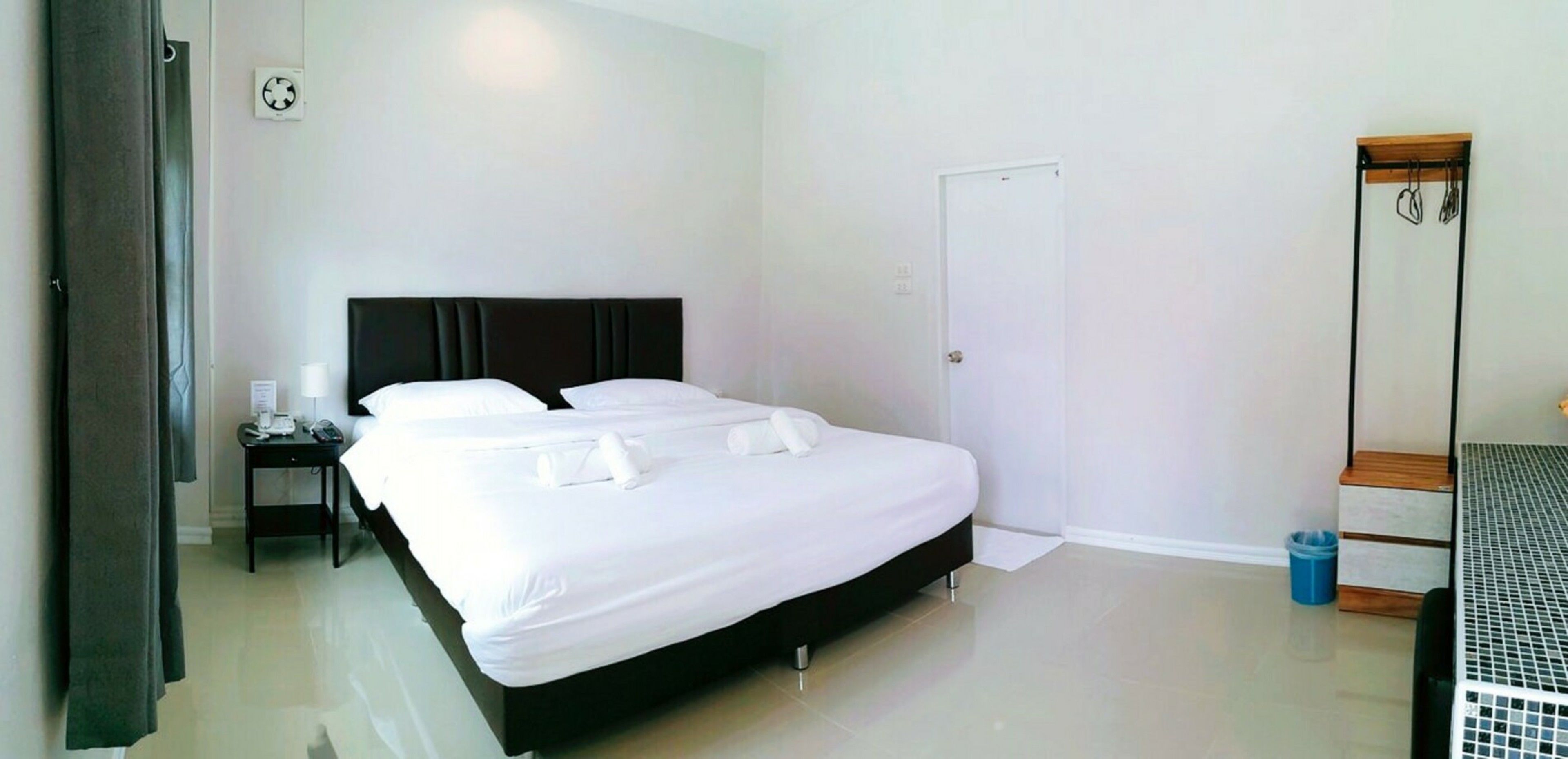 Uma cama bem arrumada com lençóis brancos, uma mesa de cabeceira com abajur e uma pequena lixeira.