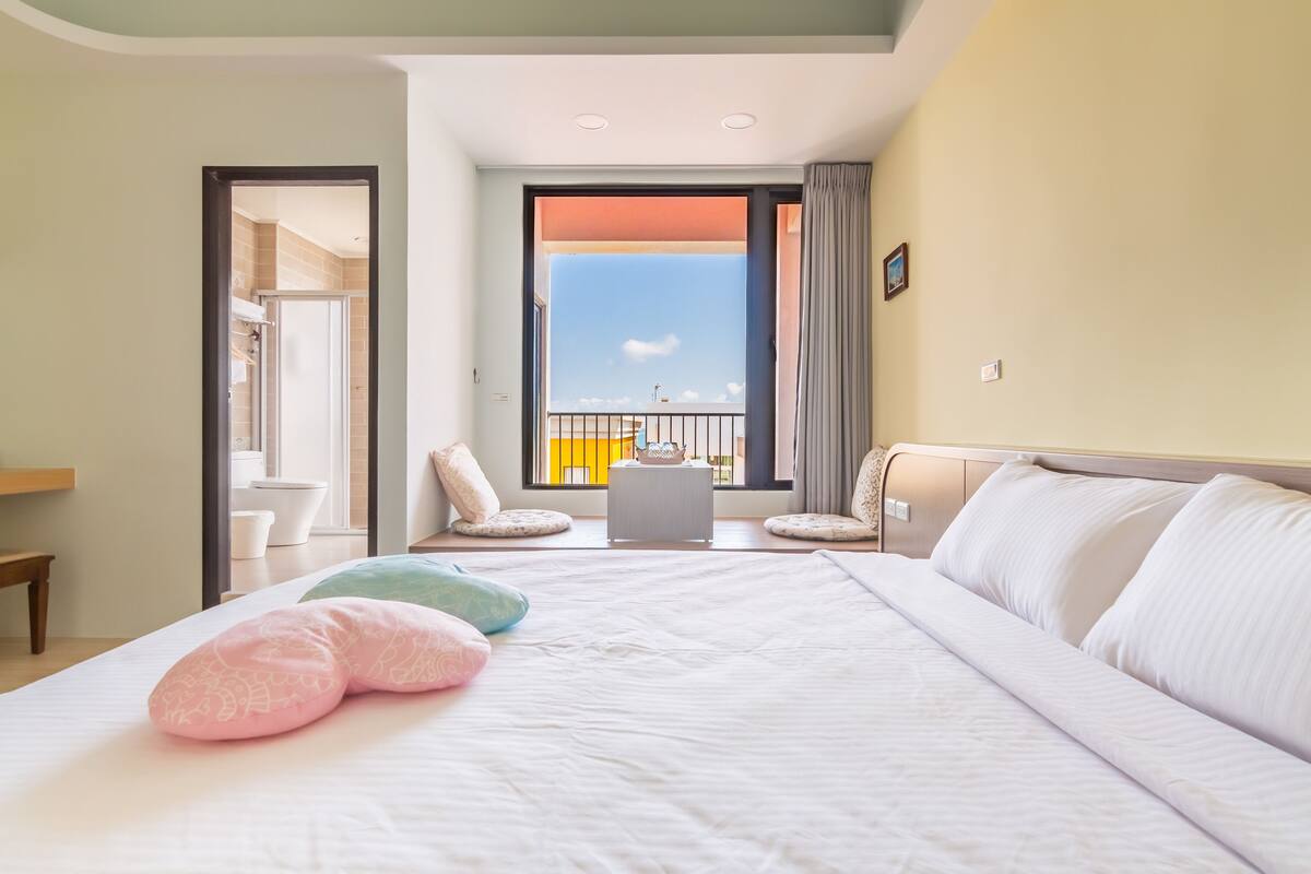 Quarto de hotel com uma cama grande, uma mesa pequena, uma cadeira e uma varanda com vista para a cidade.