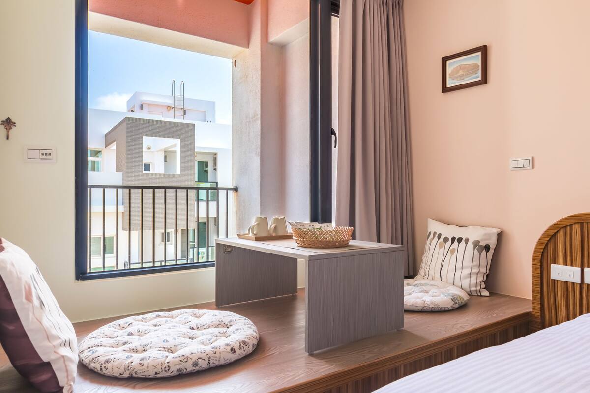 Quarto de hotel com varanda, uma pequena mesa com cesta, cama com cabeceira e janela com vista para edifícios.