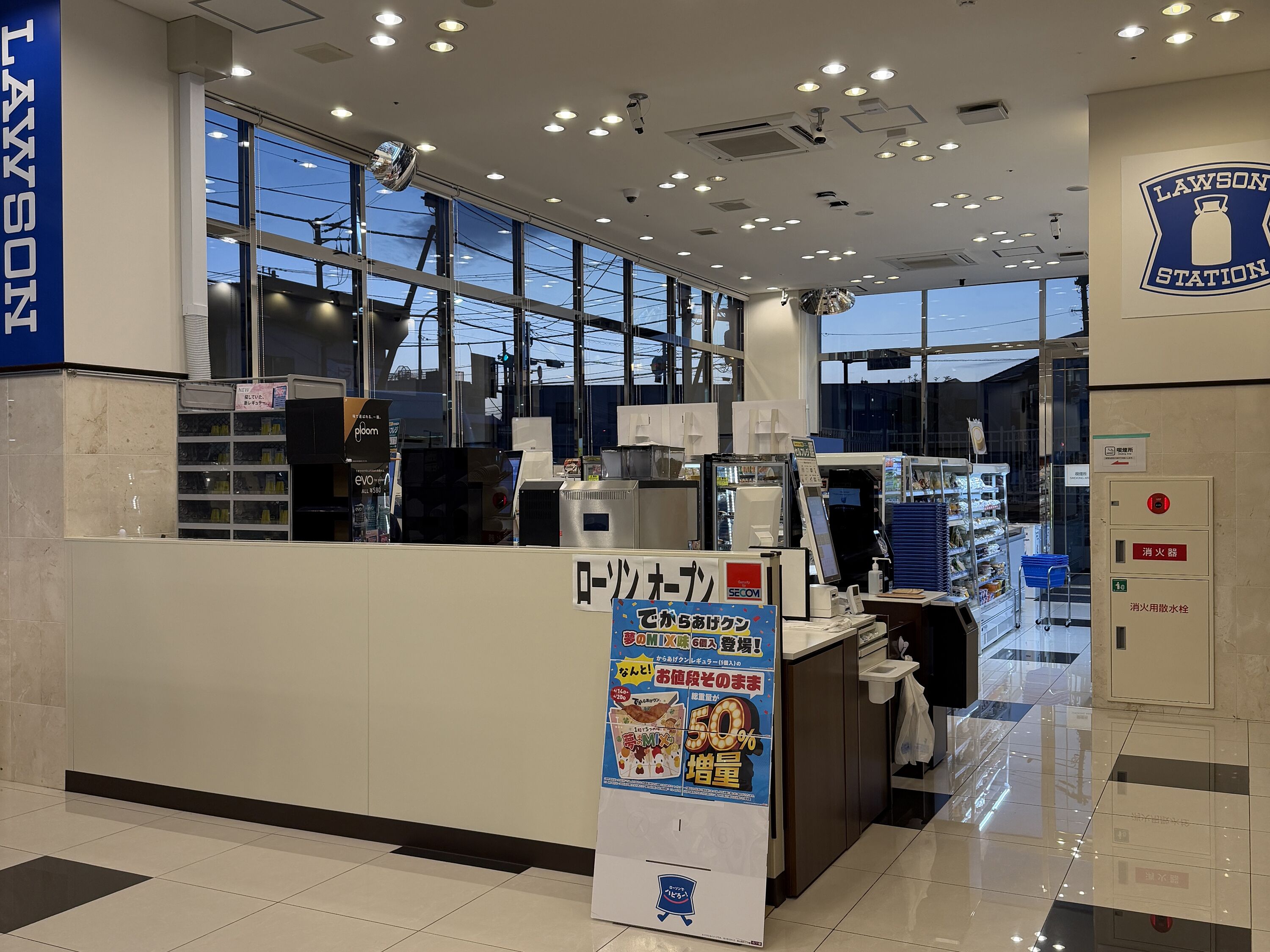 Uma loja Lawson Station com um balcão, cartazes promocionais e vista para o exterior através de grandes janelas.