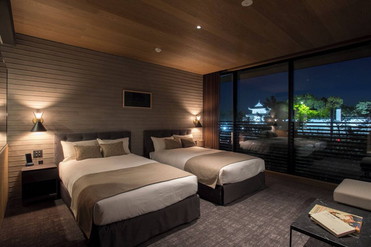 Quarto de hotel moderno com duas camas, uma janela grande com vista noturna de um edifício tradicional e uma pequena mesa com um livro.