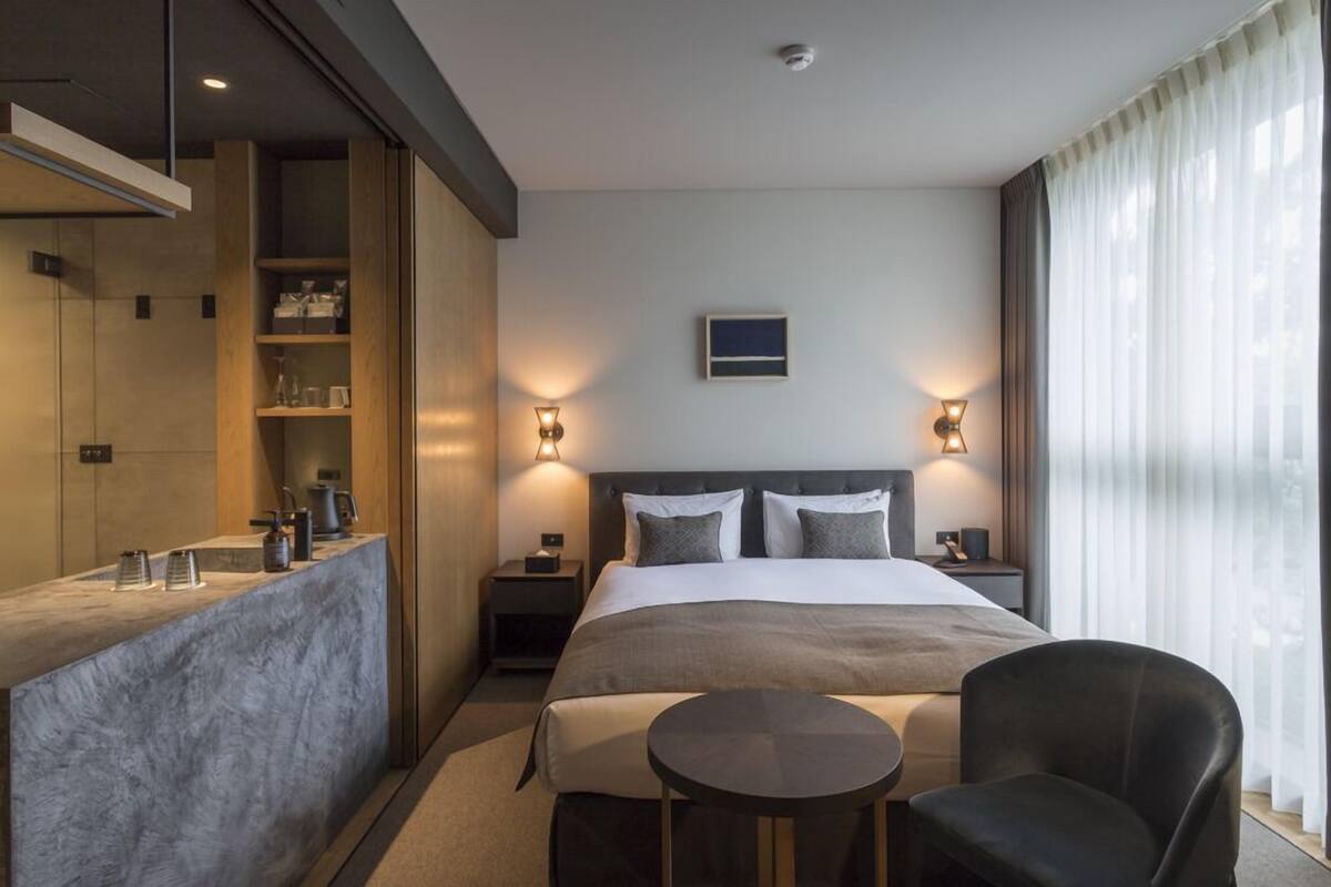 Quarto de hotel moderno com uma cama grande, mesas de cabeceira, uma mesinha redonda e um espaço de estar.