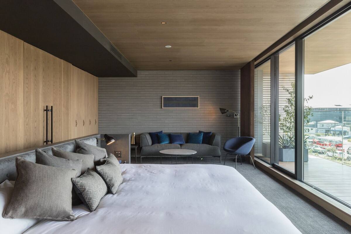 Quarto moderno com uma cama grande, um sofá e uma cadeira. Há uma janela com vista para edifícios lá fora.
